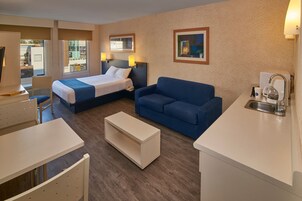 Quarto de hotel com cama, sofá azul, pequena mesa branca, cozinha compacta com pia e janela com vista para o exterior.