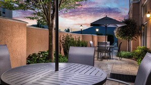 Um pátio externo com mesa redonda e cadeiras, rodeado por um muro baixo e plantas.