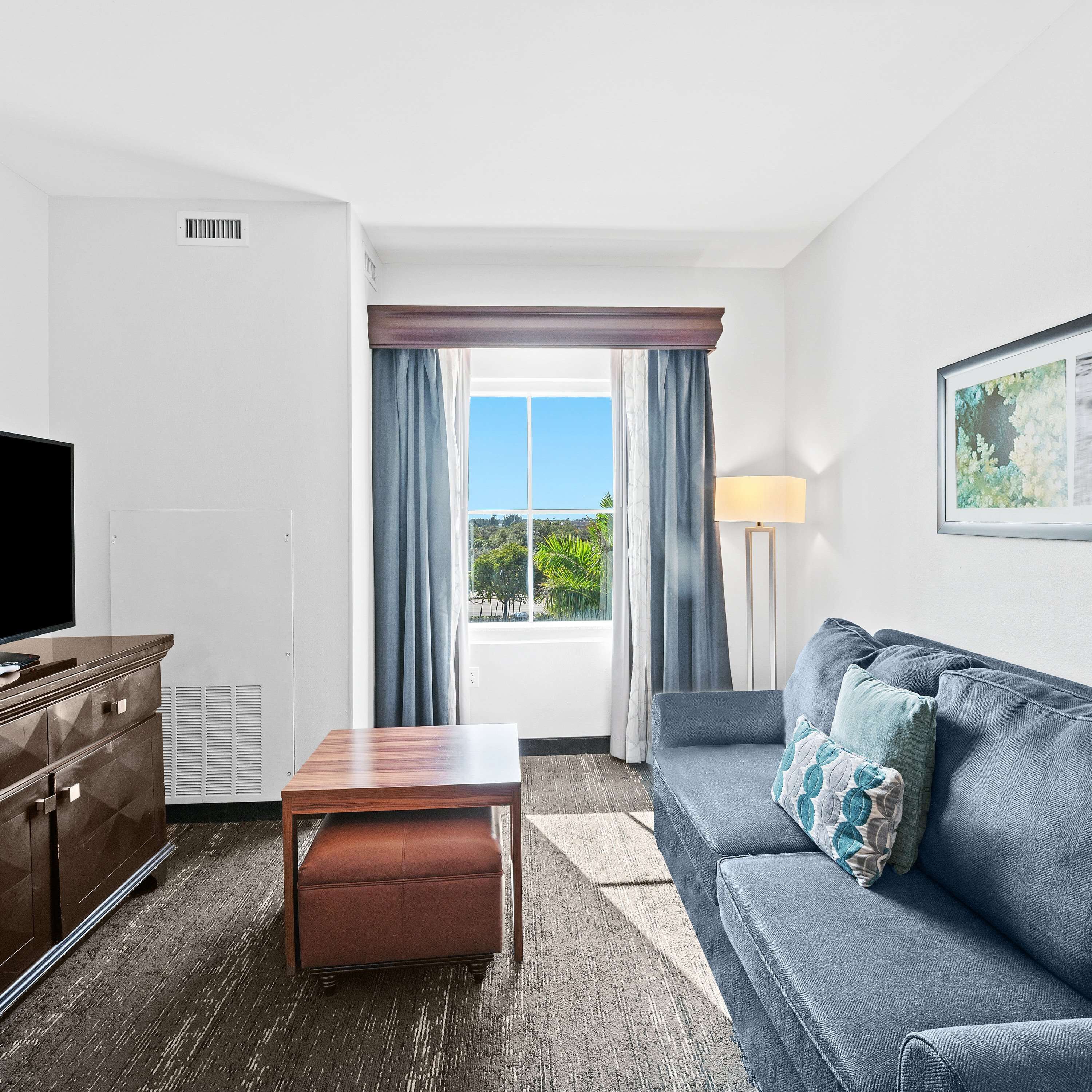 Quarto de hotel moderno com uma grande janela, TV de tela plana, mesa de centro de madeira, sofá e abajur.
