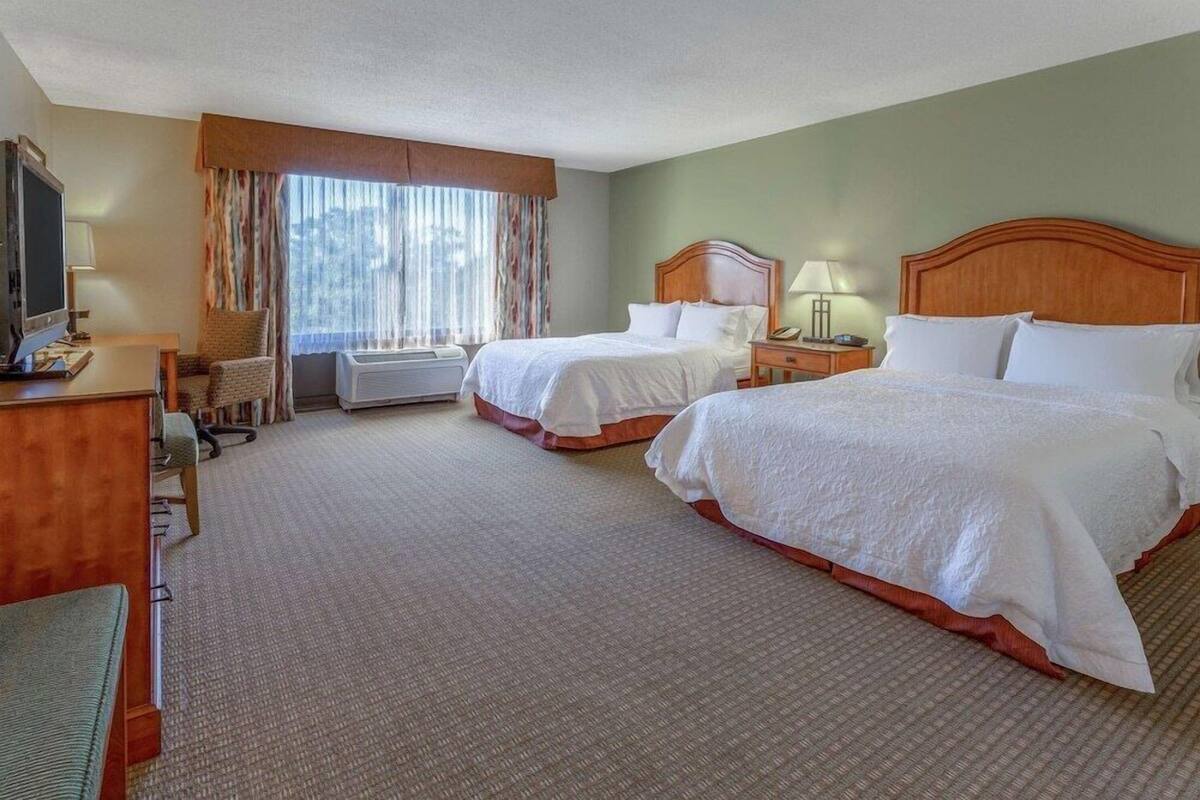 Quarto de hotel com duas camas, uma escrivaninha, uma cadeira, uma televisão e uma janela com cortinas.