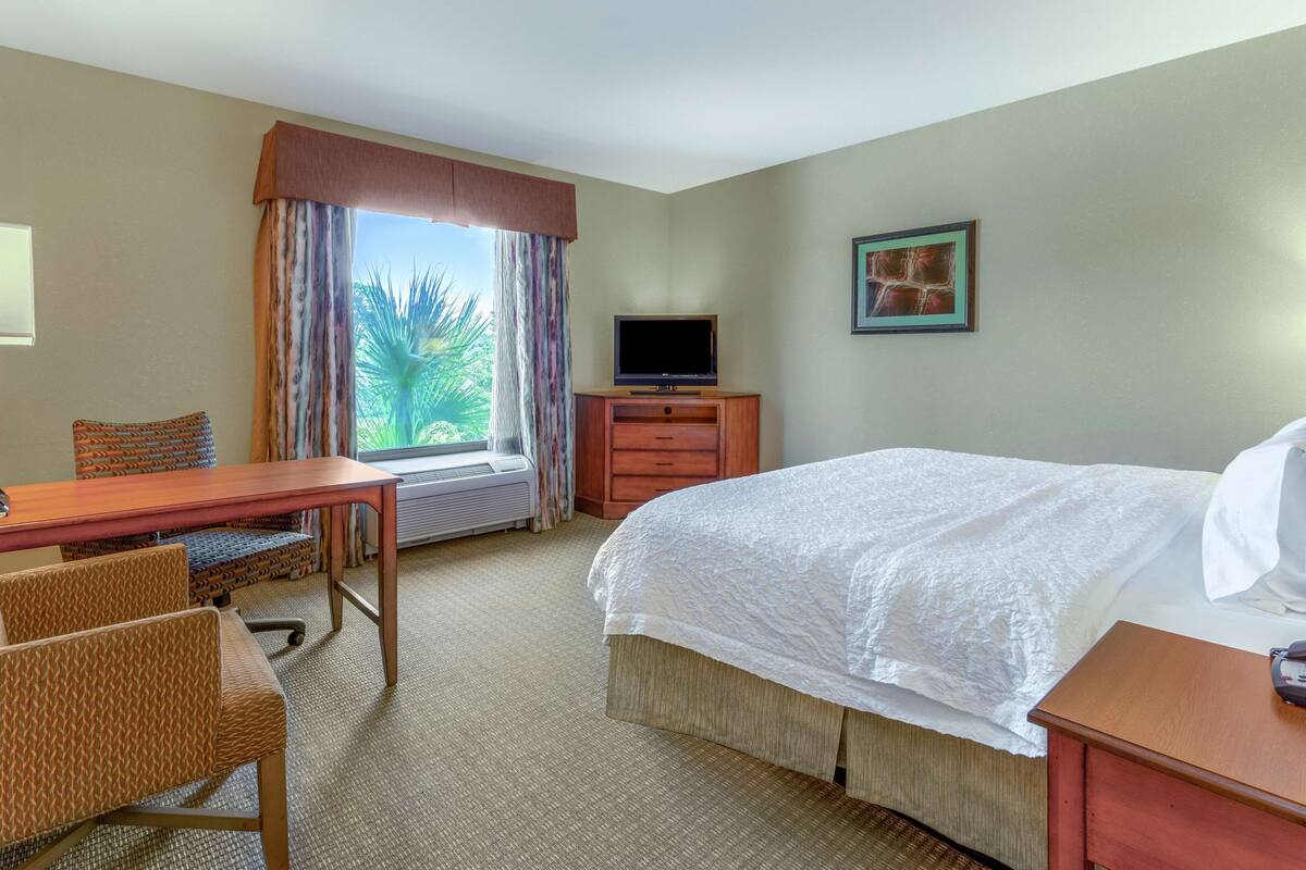 Quarto de hotel com cama, escrivaninha, cadeira, televisão e janela com cortinas.