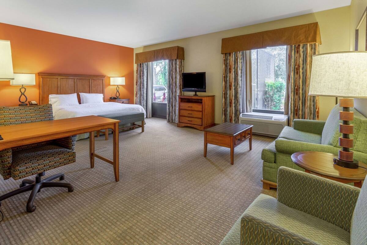 Quarto de hotel com cama, escrivaninha, cadeira, sofá, televisão e cortinas.