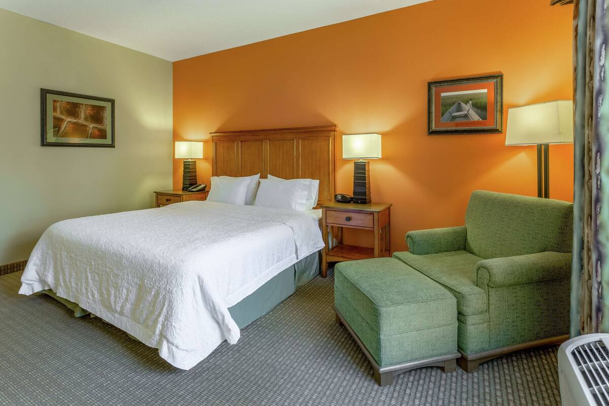 Quarto de hotel com uma cama, duas mesas de cabeceira com luminárias, uma poltrona verde e um quadro na parede.