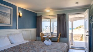 Quarto de hotel com cama, poltrona, mesa, janela com cortinas e porta para uma praia.