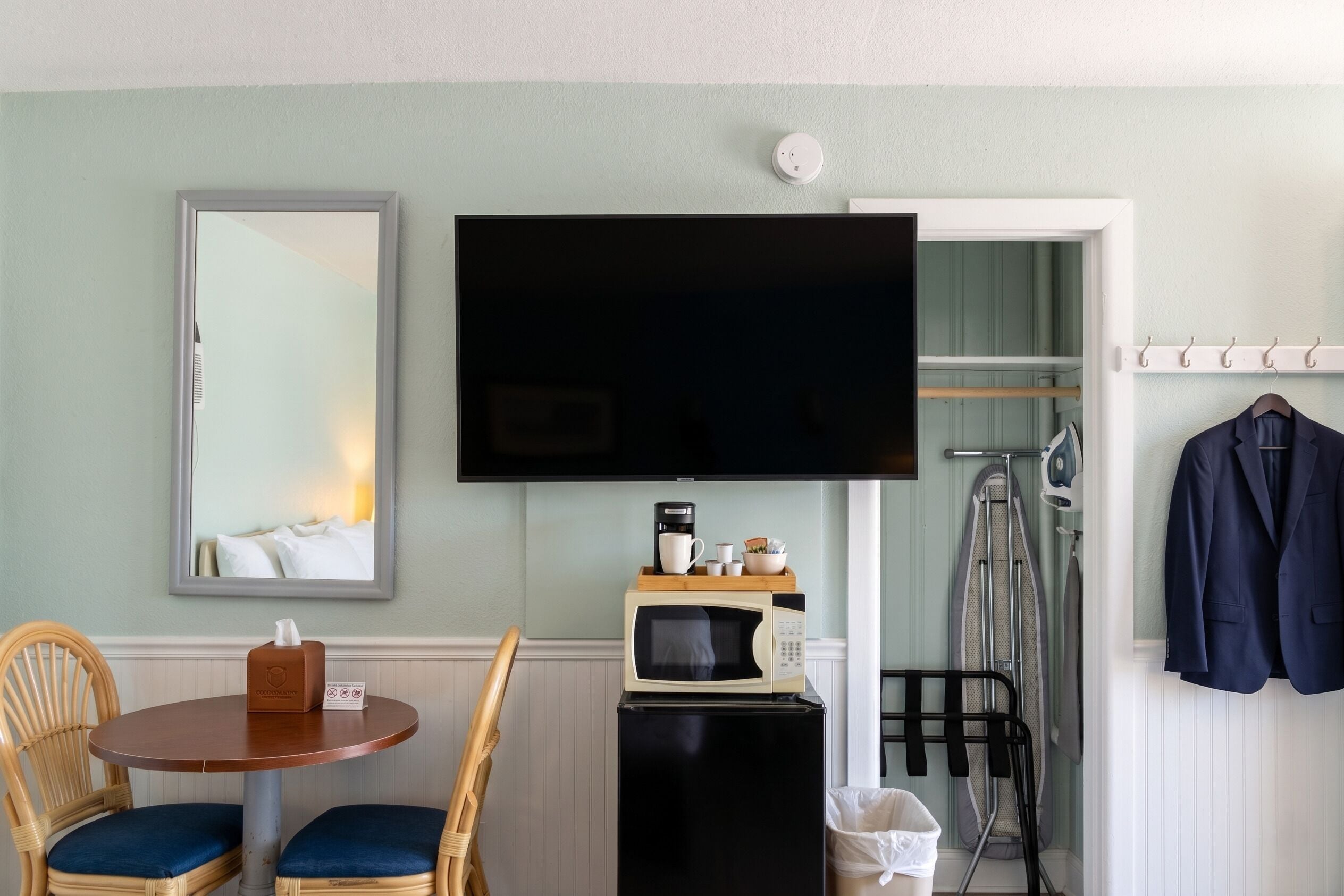 Quarto de hotel com televisão de tela plana, micro-ondas, mini-geladeira, mesa com cadeiras, espelho, tábua de passar roupa e cabideiro.