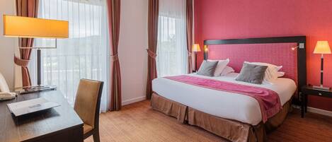Quarto de hotel com cama, luminárias de cabeceira, escrivaninha com cadeira e janela com cortinas.