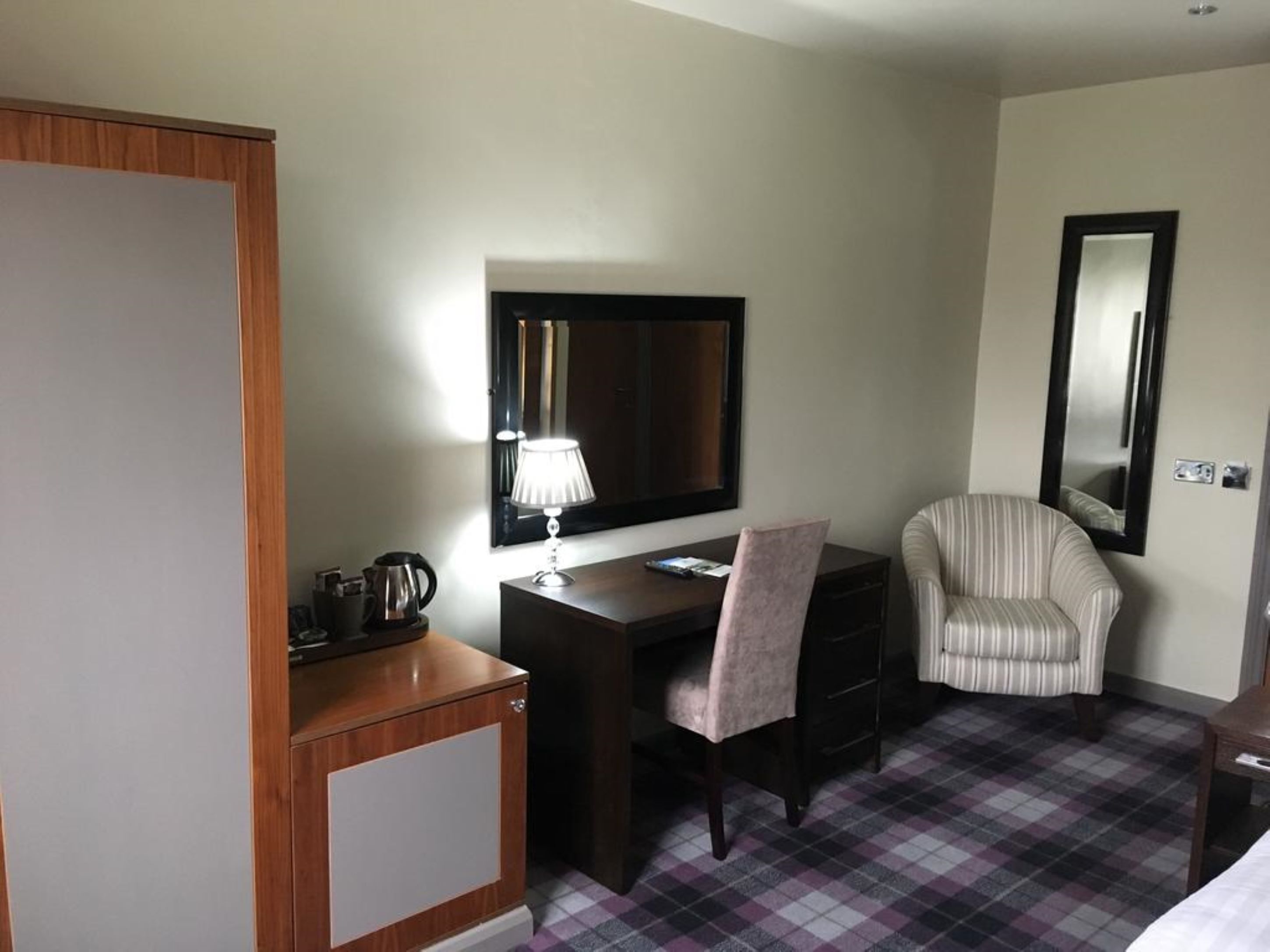 Quarto de hotel com um armário de madeira, uma escrivaninha com abajur, uma poltrona listrada e um espelho grande na parede.