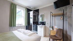 Quarto de hotel com cama, escrivaninha, cadeira, chuveiro e televisão.