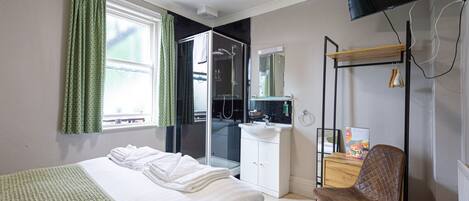 Quarto de hotel com cama, escrivaninha, cadeira, chuveiro e televisão.