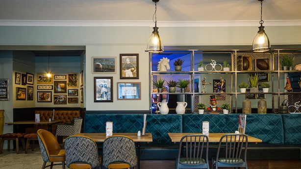 Restaurante com área de bar, mesas e cadeiras, e parede decorada com fotos emolduradas e plantas.