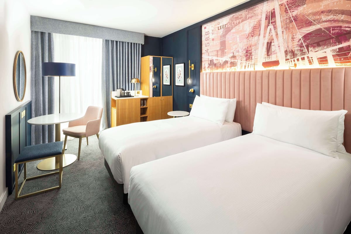 Quarto de hotel com duas camas, uma escrivaninha, uma cadeira, um guarda-roupa e uma janela grande com cortinas.