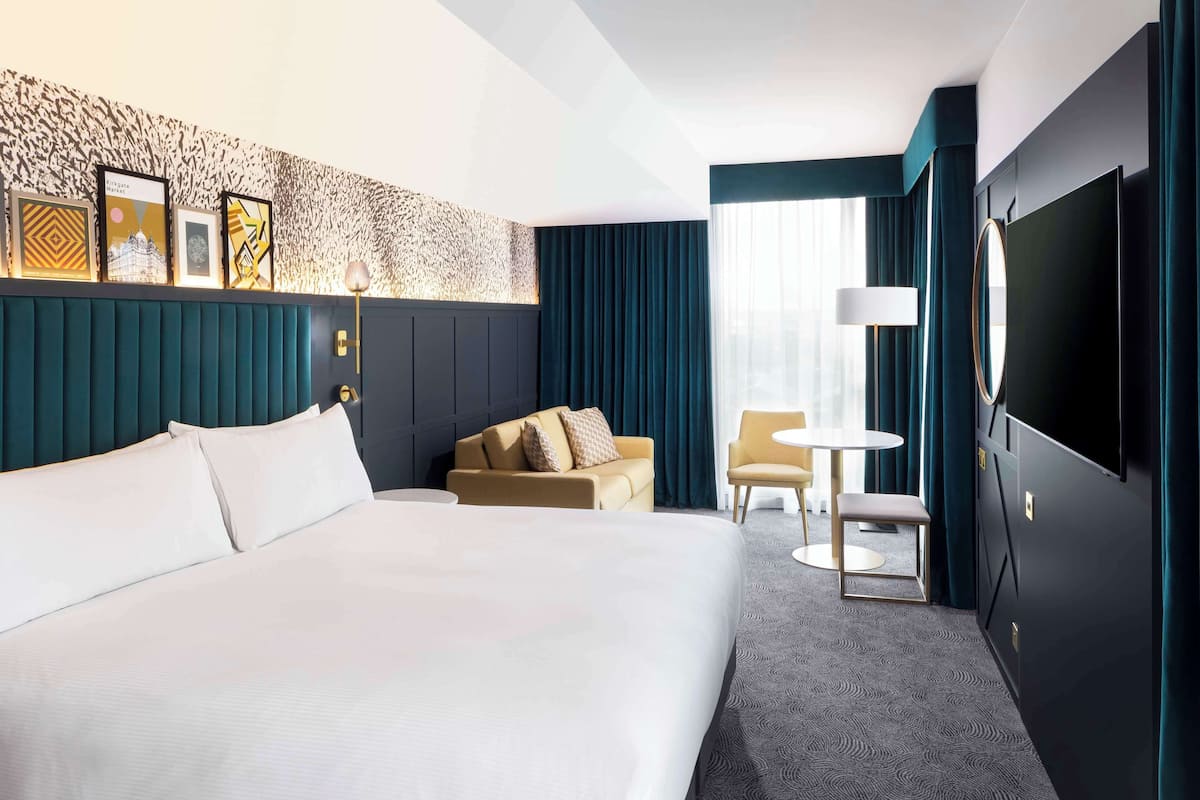Quarto de hotel com cama grande, sofá, mesa pequena, cadeira, televisão e obras de arte na parede.