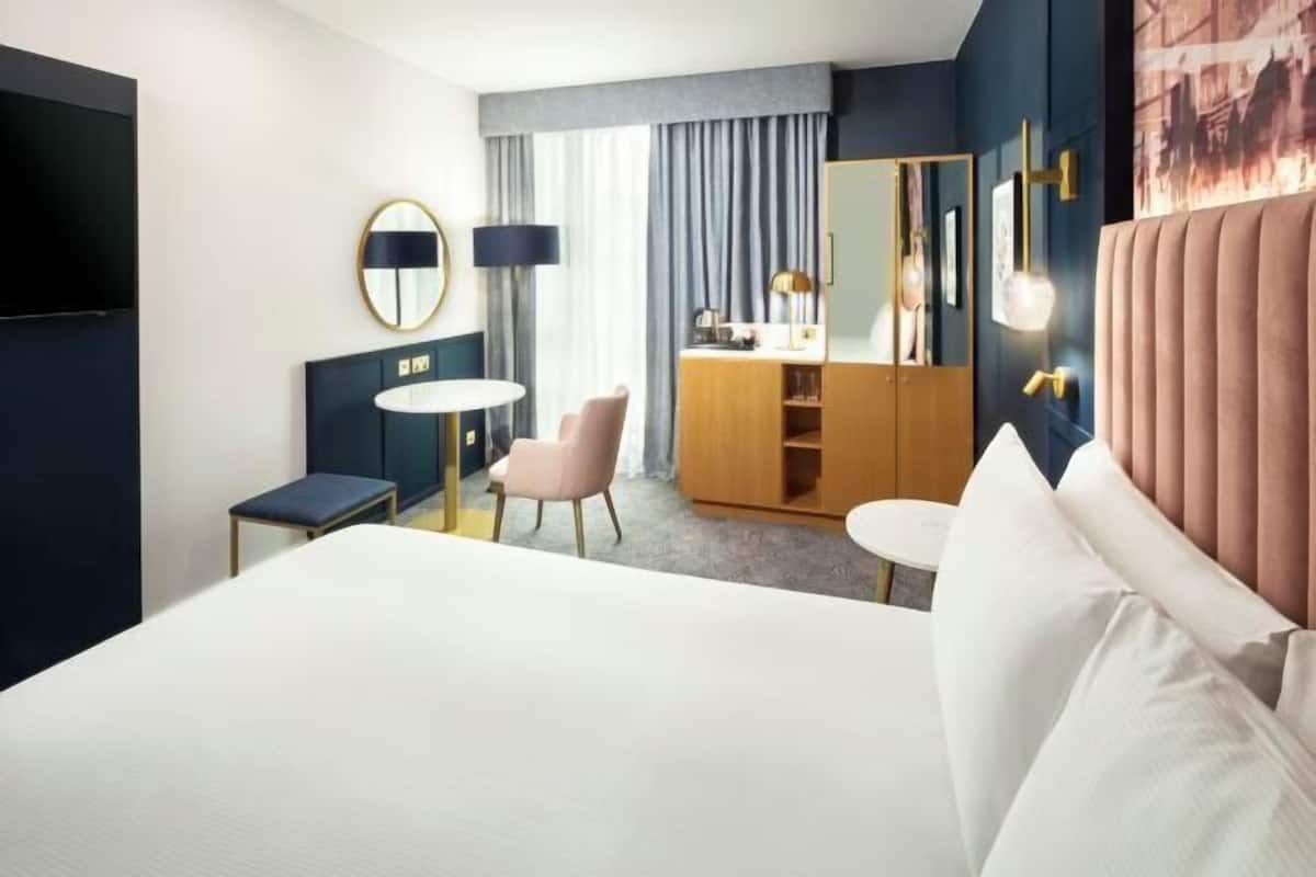 Quarto de hotel com uma cama grande, uma escrivaninha, uma cadeira, um espelho e uma televisão.