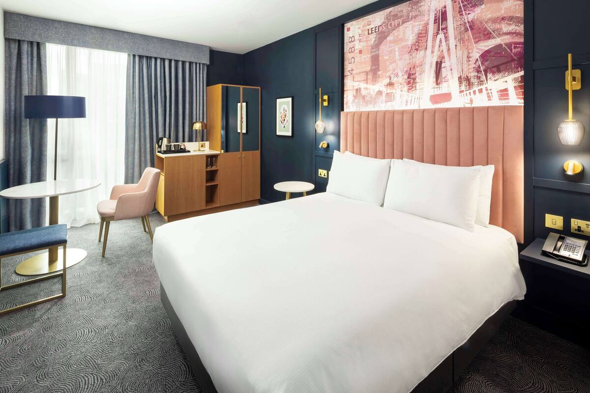 Quarto de hotel com uma cama grande, uma mesa, uma cadeira, uma mesinha e um telefone fixado na parede.