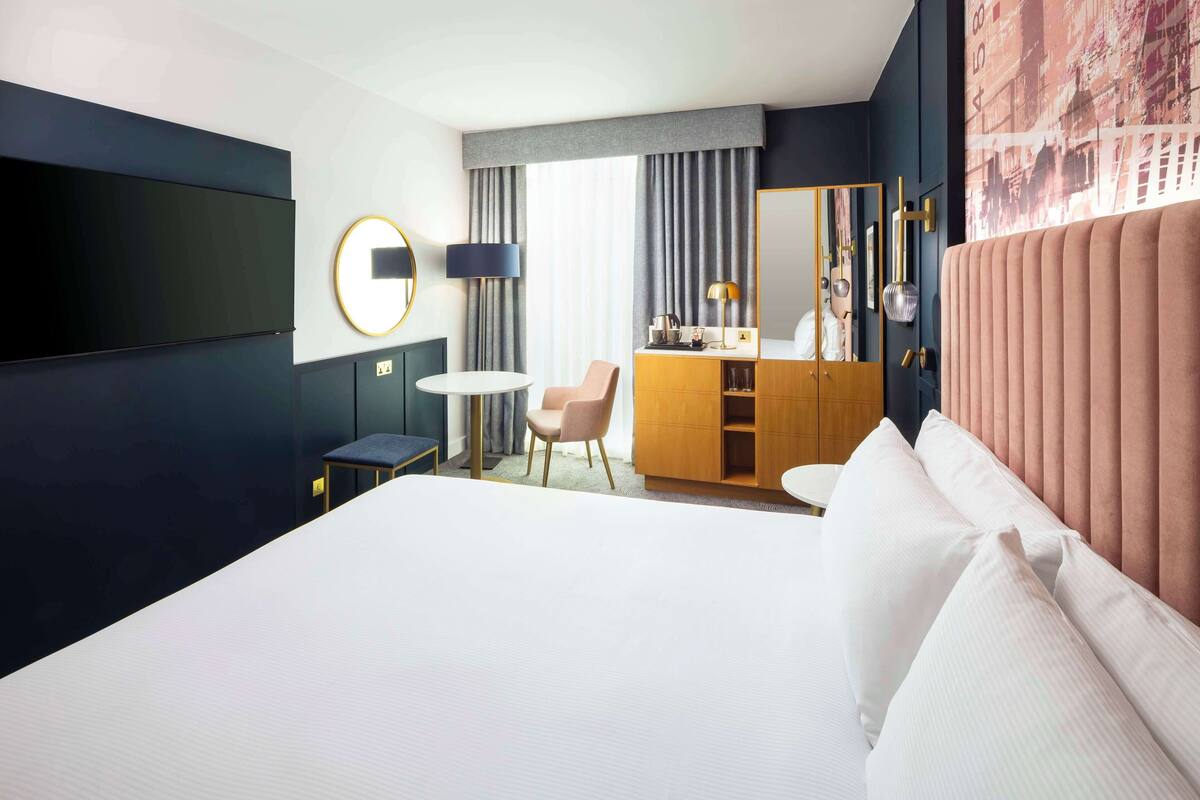 Quarto de hotel com uma cama grande, uma mesa de trabalho com cadeira, uma televisão fixada na parede e um espelho amplo.