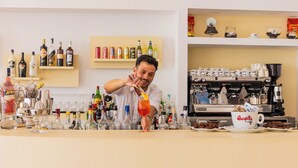 Uma área de bar com diversas garrafas e copos, uma máquina de café e um balcão onde alguém prepara uma bebida.