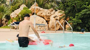 Uma piscina com uma pessoa salpicando água, rodeada por palmeiras e um cenário rochoso.