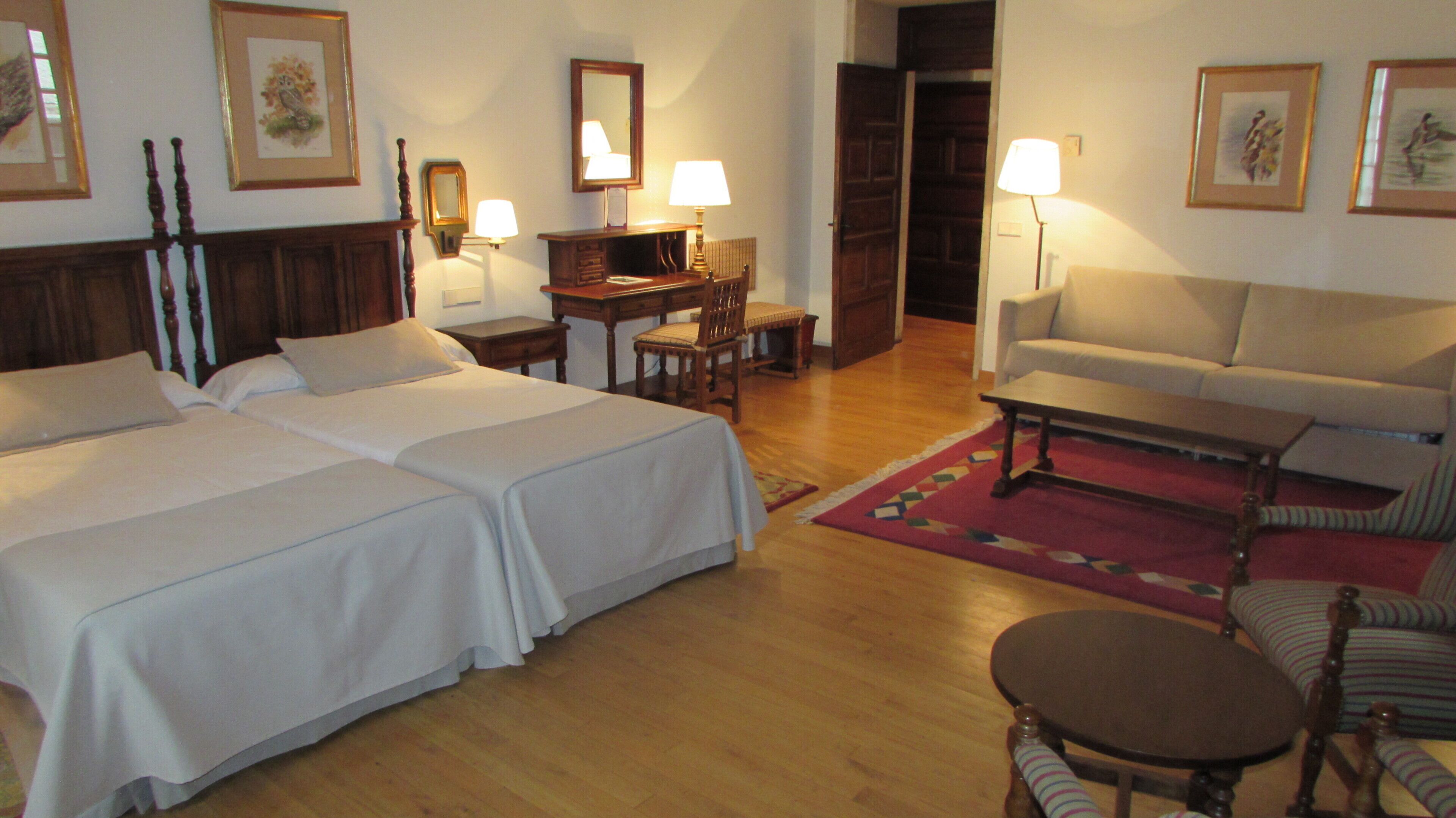 Quarto de hotel com duas camas, um sofá, uma mesa pequena e uma cadeira. Há luminárias nas paredes e uma porta ao fundo.