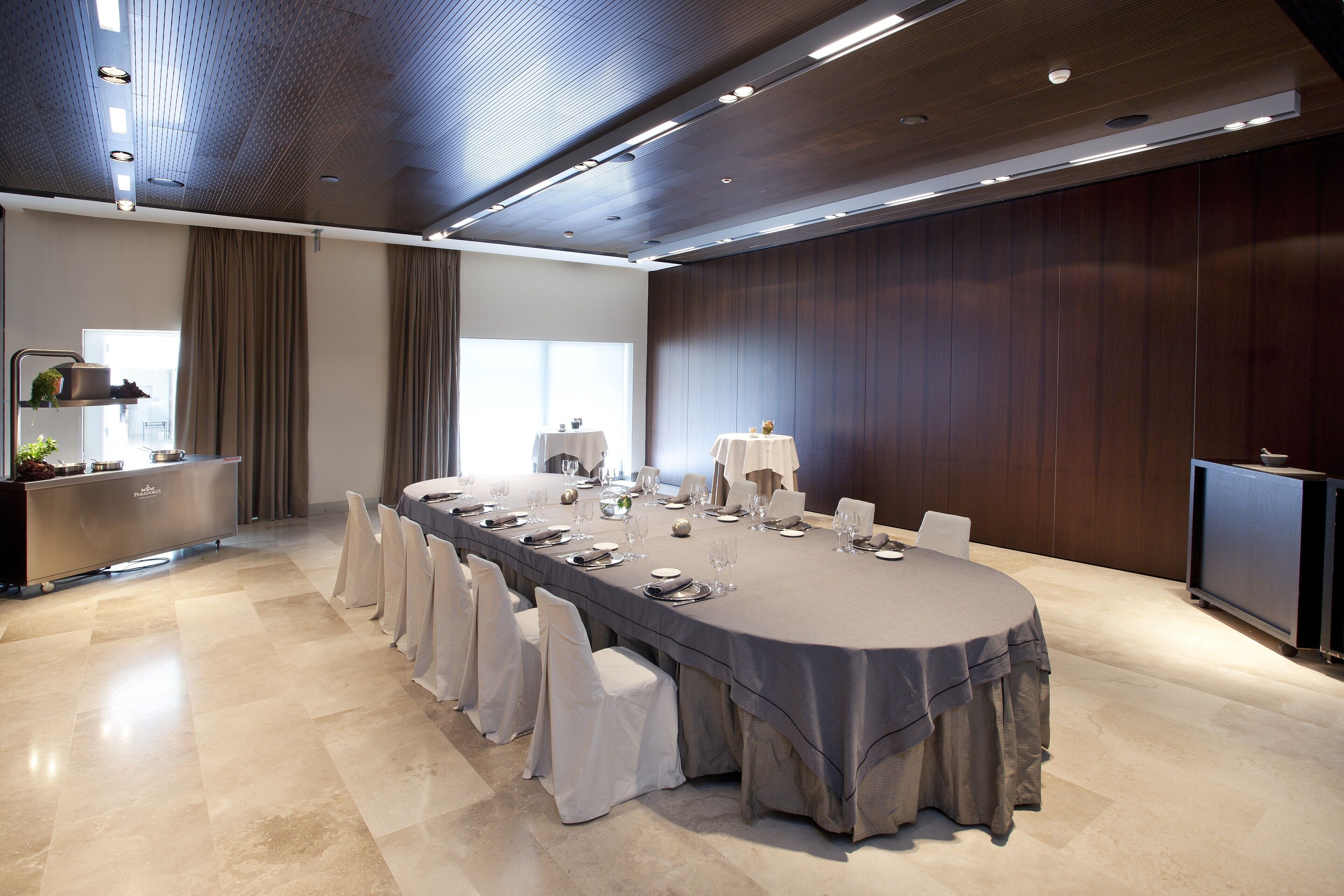 Uma sala de conferências com uma mesa comprida preparada para uma reunião, um espaço para buffet e uma televisão sobre um móvel.