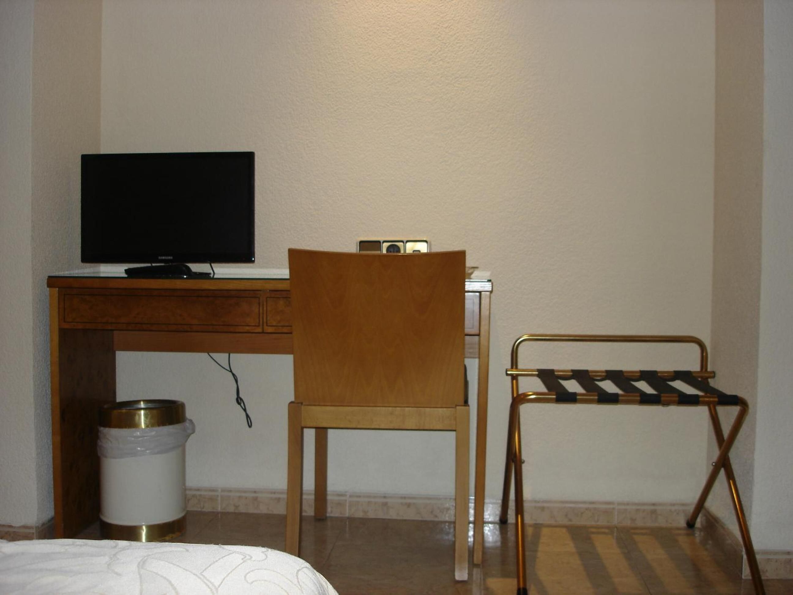 Quarto de hotel com escrivaninha de madeira, televisão, cadeira, porta-bagagem de metal e lixeira.
