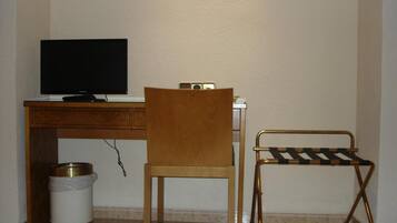 Quarto de hotel com escrivaninha de madeira, televisão, cadeira, porta-bagagem de metal e lixeira.