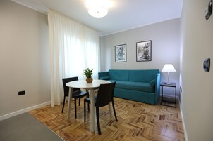 Uma sala de estar moderna com um sofá verde-água, uma mesa de jantar redonda com cadeiras, uma mesinha de apoio com abajur e duas fotografias emolduradas na parede.