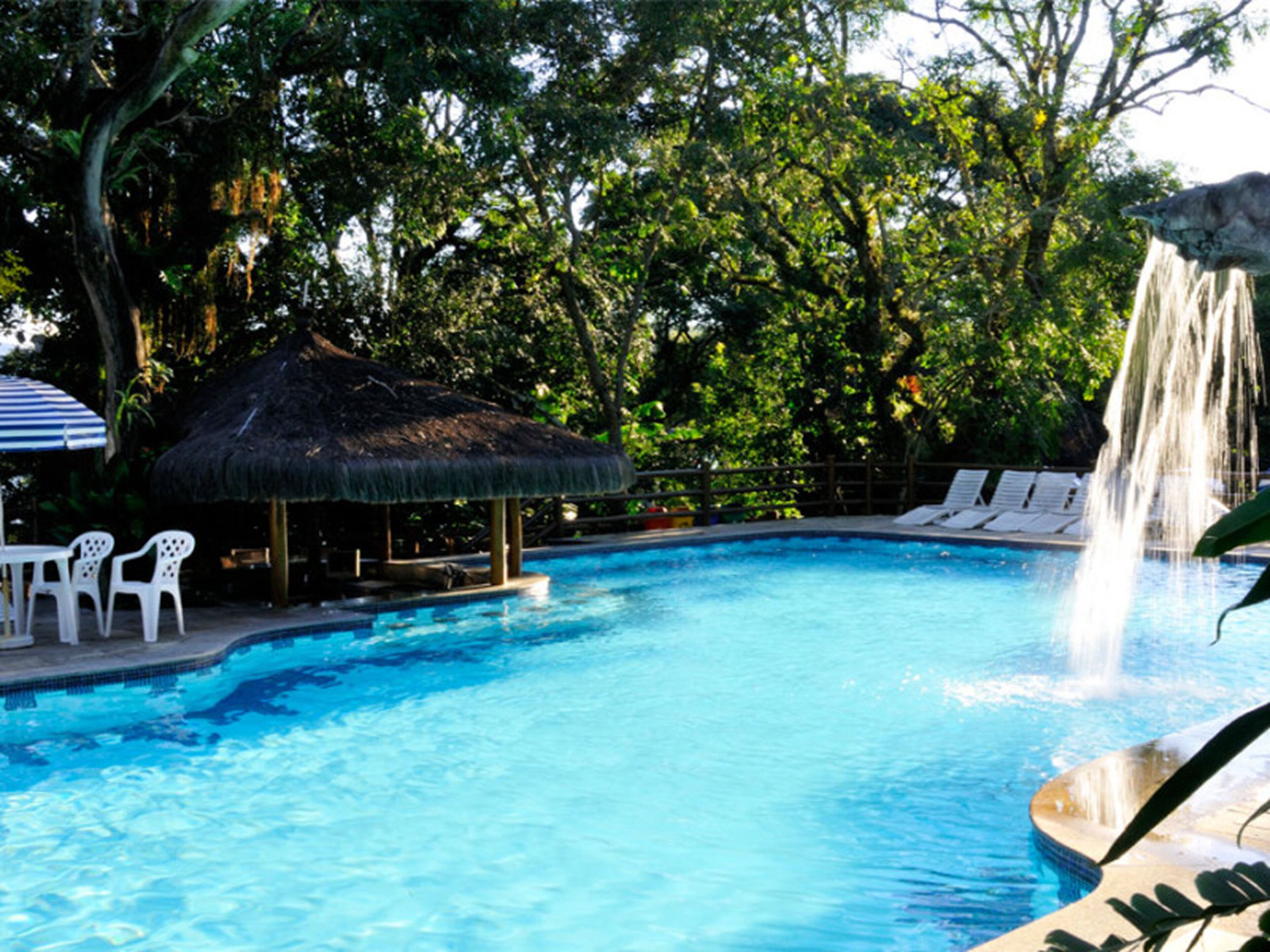 Uma piscina com cachoeira artificial, rodeada por espreguiçadeiras e uma construção com telhado de palha.