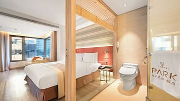 Quarto de hotel com uma cama grande, uma cama menor, banheiro com vaso sanitário e vista para o exterior através de uma janela ampla.
