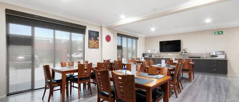 Uma sala de jantar de hotel com mesas e cadeiras de madeira, uma cozinha ao fundo e uma grande janela com vista para carros estacionados.
