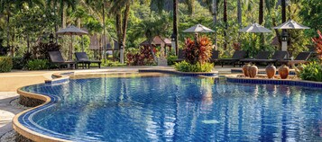 Uma piscina com espreguiçadeiras e guarda-sóis, cercada por plantas tropicais e um gazebo.