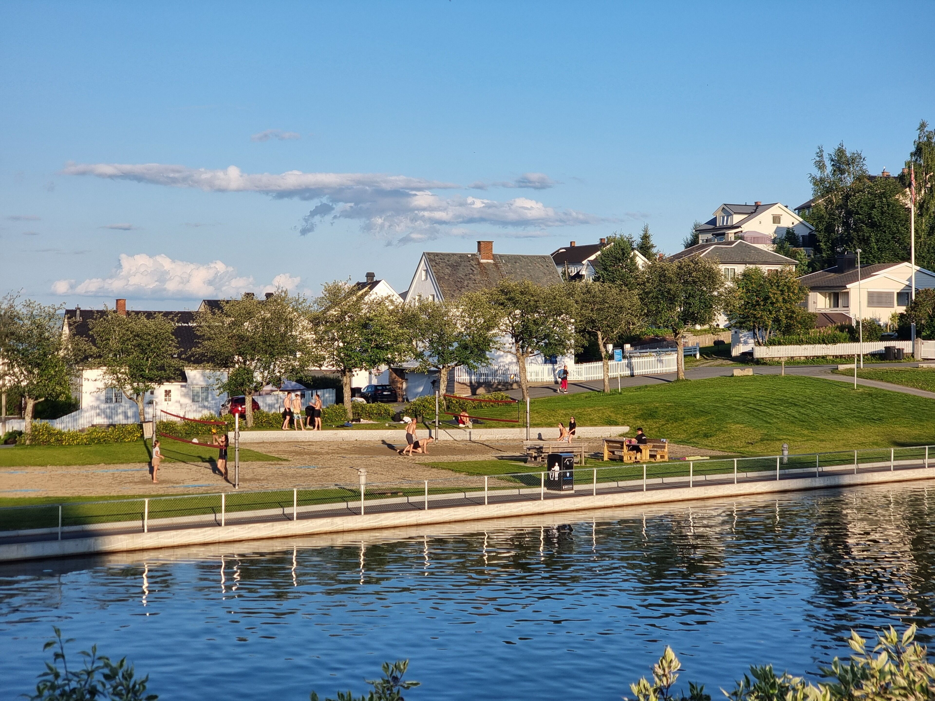 Um parque à beira do lago com quadra de voleibol, bancos e uma trilha para caminhada.