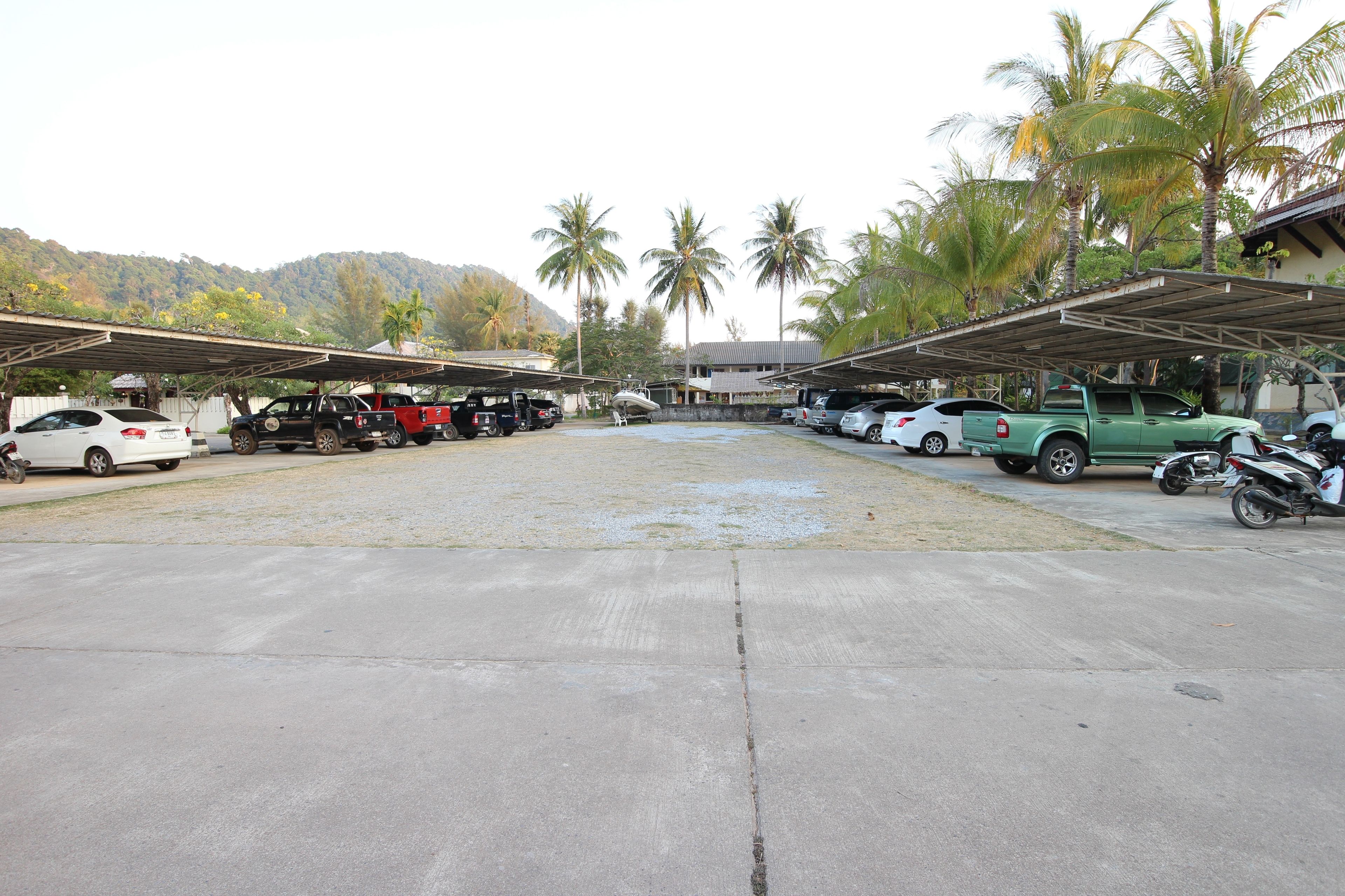 Uma área de estacionamento com vários veículos, incluindo carros e motocicletas, sob estruturas cobertas e palmeiras ao fundo.
