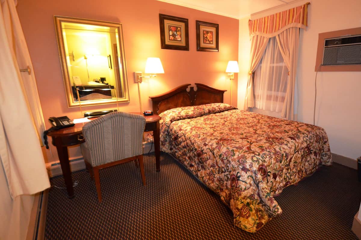 Quarto de hotel com uma cama, uma escrivaninha com telefone, duas luminárias e um espelho.