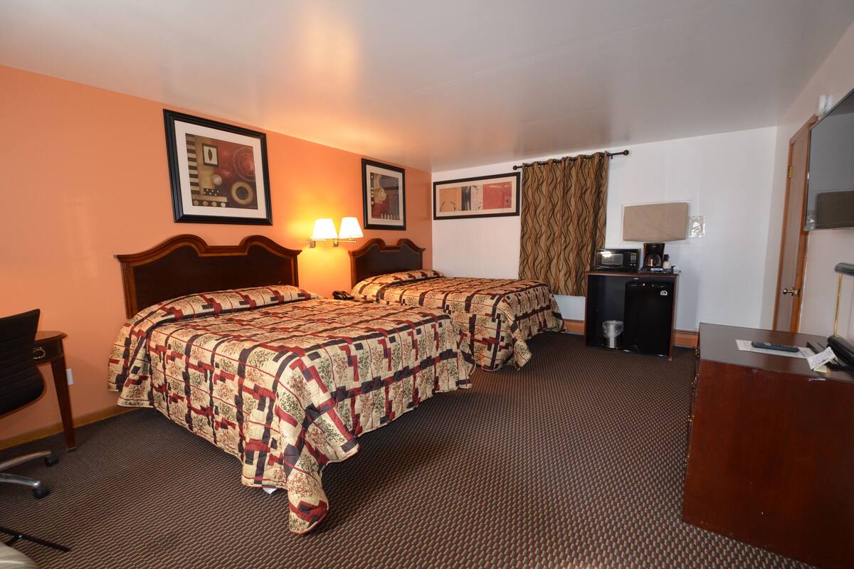 Quarto de hotel com duas camas, uma mesa, uma cadeira e uma luminária de mesa.