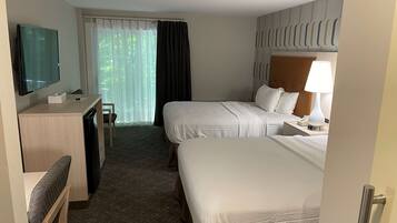 Quarto de hotel com duas camas, uma escrivaninha com televisão, uma cadeira, uma janela com cortinas e um abajur.