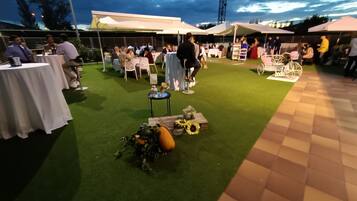 Evento ao ar livre com mesas e cadeiras sob toldos brancos, área gramada e um caminho de azulejos.