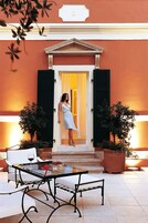 Uma mulher de vestido branco está em frente à entrada de um edifício com porta amarela e persianas pretas. Há uma mesa com tampo de vidro e duas cadeiras em frente à porta.