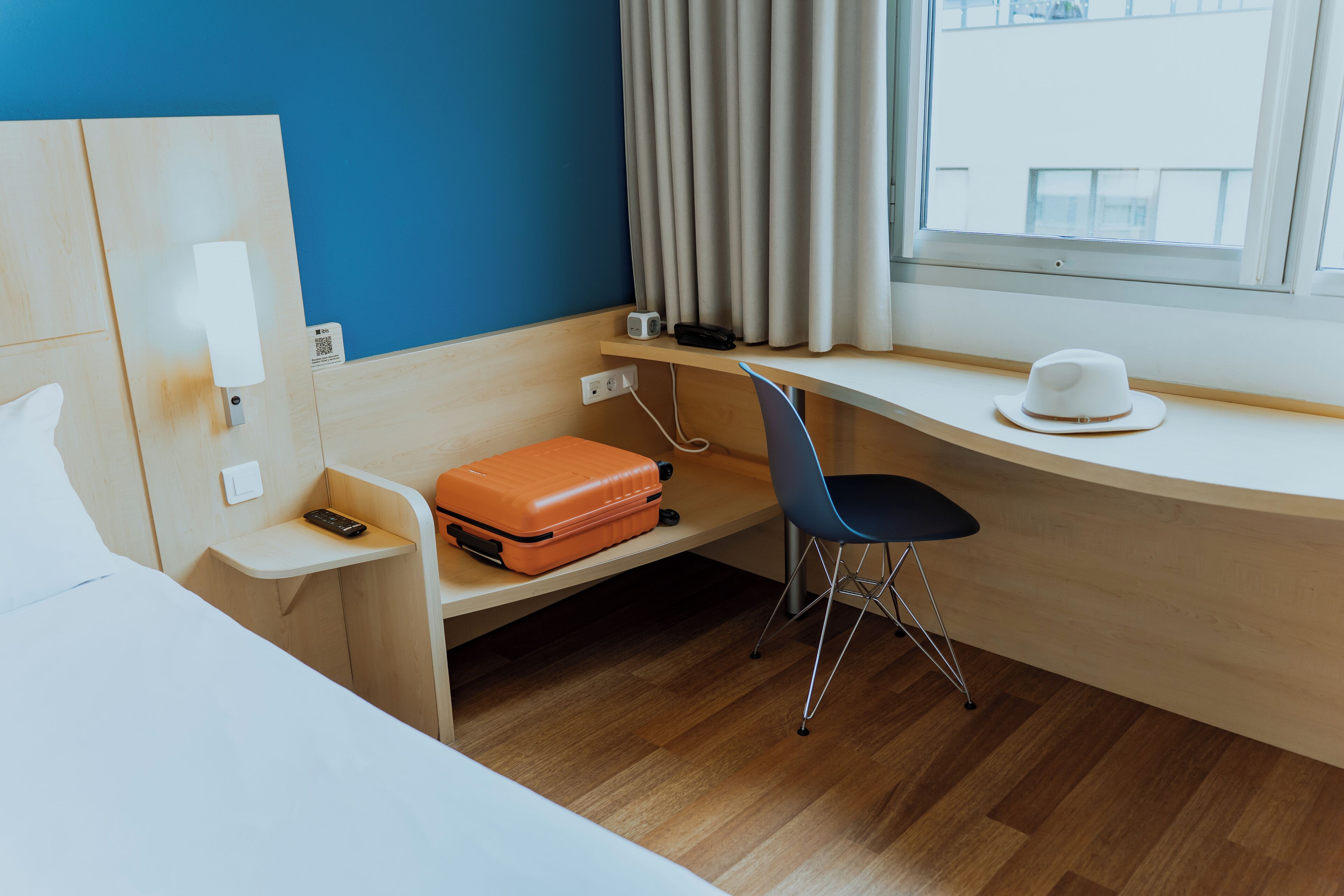 Quarto de hotel com cama, escrivaninha com cadeira, mala e janela com cortinas.