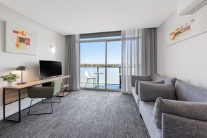 Quarto de hotel moderno com sofá cinzento, secretária com televisão de ecrã plano, pequena mesa com cadeira e varanda com vista para a cidade.