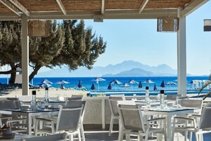 Um restaurante à beira-mar com mesas, cadeiras e guarda-sóis ao ar livre, com vista para o mar e as montanhas.