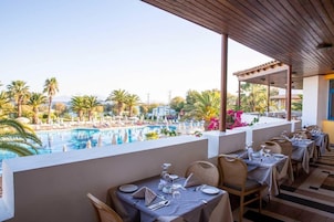 Restaurante com área externa voltada para uma piscina, cercado por palmeiras e céu claro.