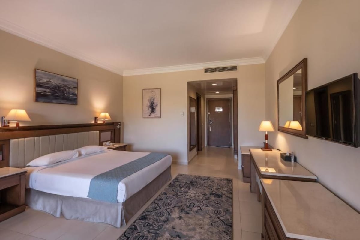 Quarto de hotel com uma cama, mesas de cabeceira, uma escrivaninha, um espelho e uma porta que dá para outro cômodo.