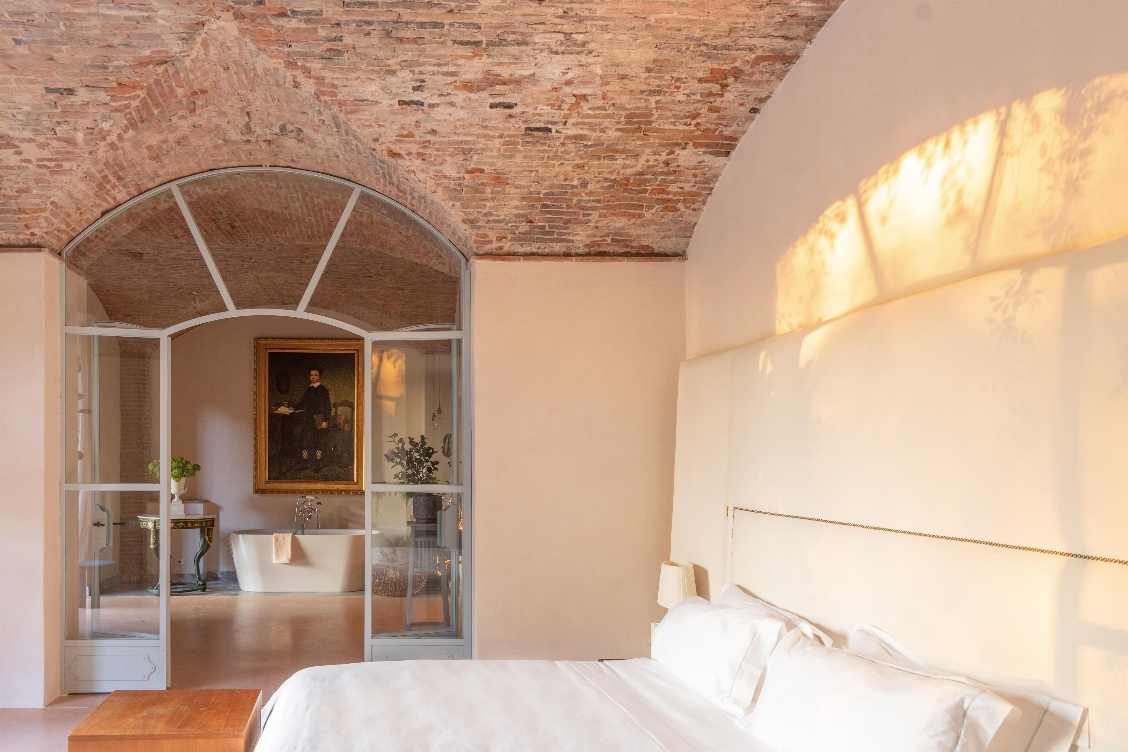 Quarto com teto de tijolos, uma cama grande, um quadro e vista através de uma porta em arco.