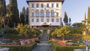 Um edifício grande e com vários andares, com fachada simétrica, entrada central com degraus e um jardim formal com sebes bem aparadas e plantas em vasos.