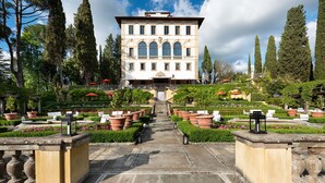 Um edifício imponente com fachada simétrica, entrada central e um jardim formal com caminhos e plantas em vasos.