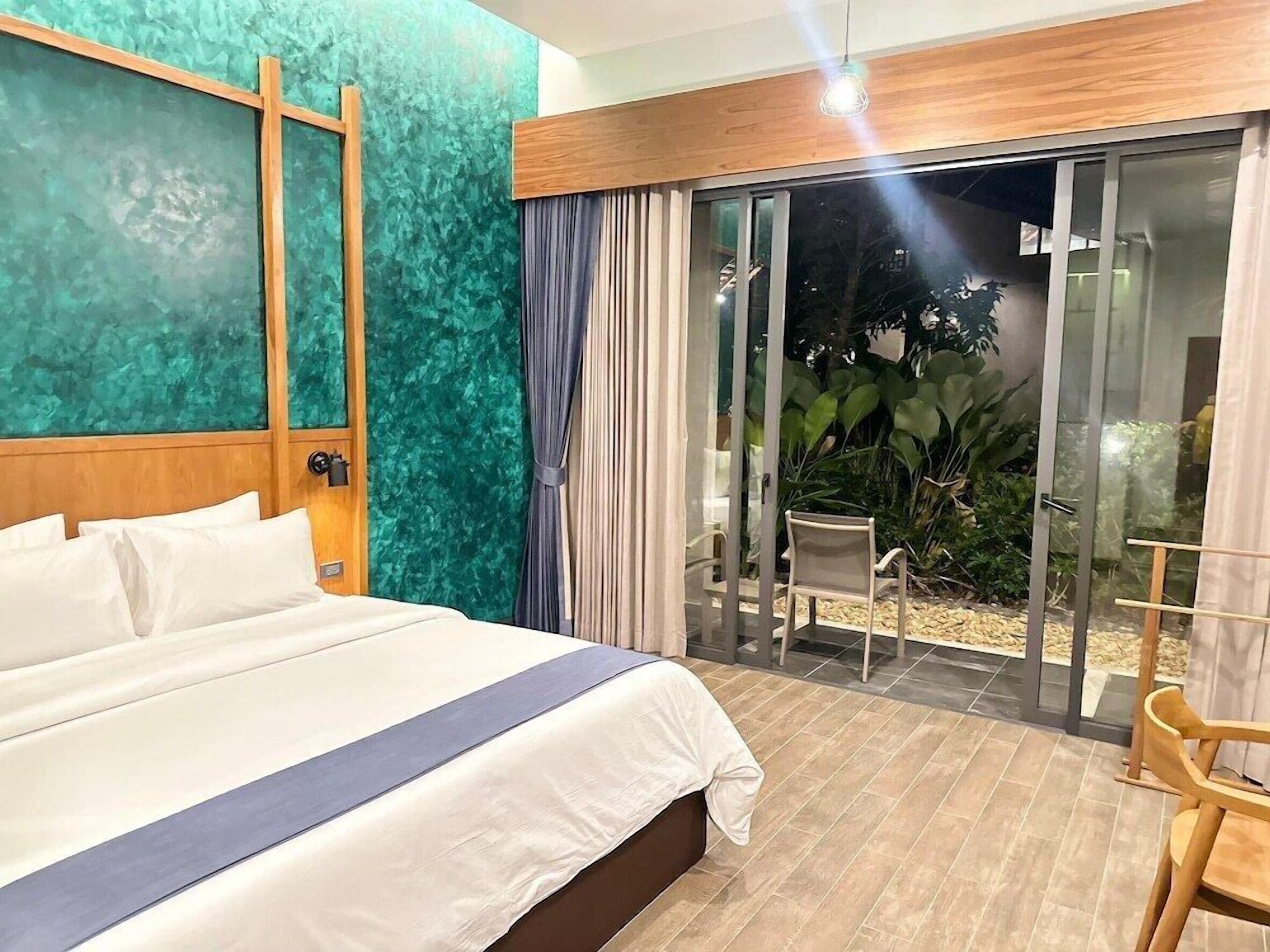 Quarto de hotel moderno com uma cama grande, cabeceira de madeira, porta de vidro deslizante para área externa com plantas e uma cadeira.