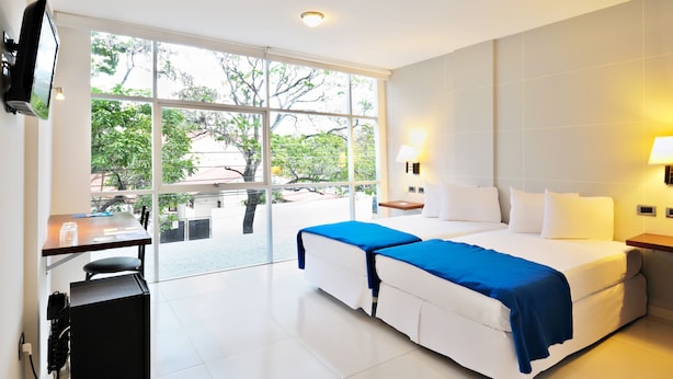 Quarto de hotel com uma janela grande, uma escrivaninha com cadeira, cama com roupa de cama branca e detalhes azuis, e uma TV fixada na parede.