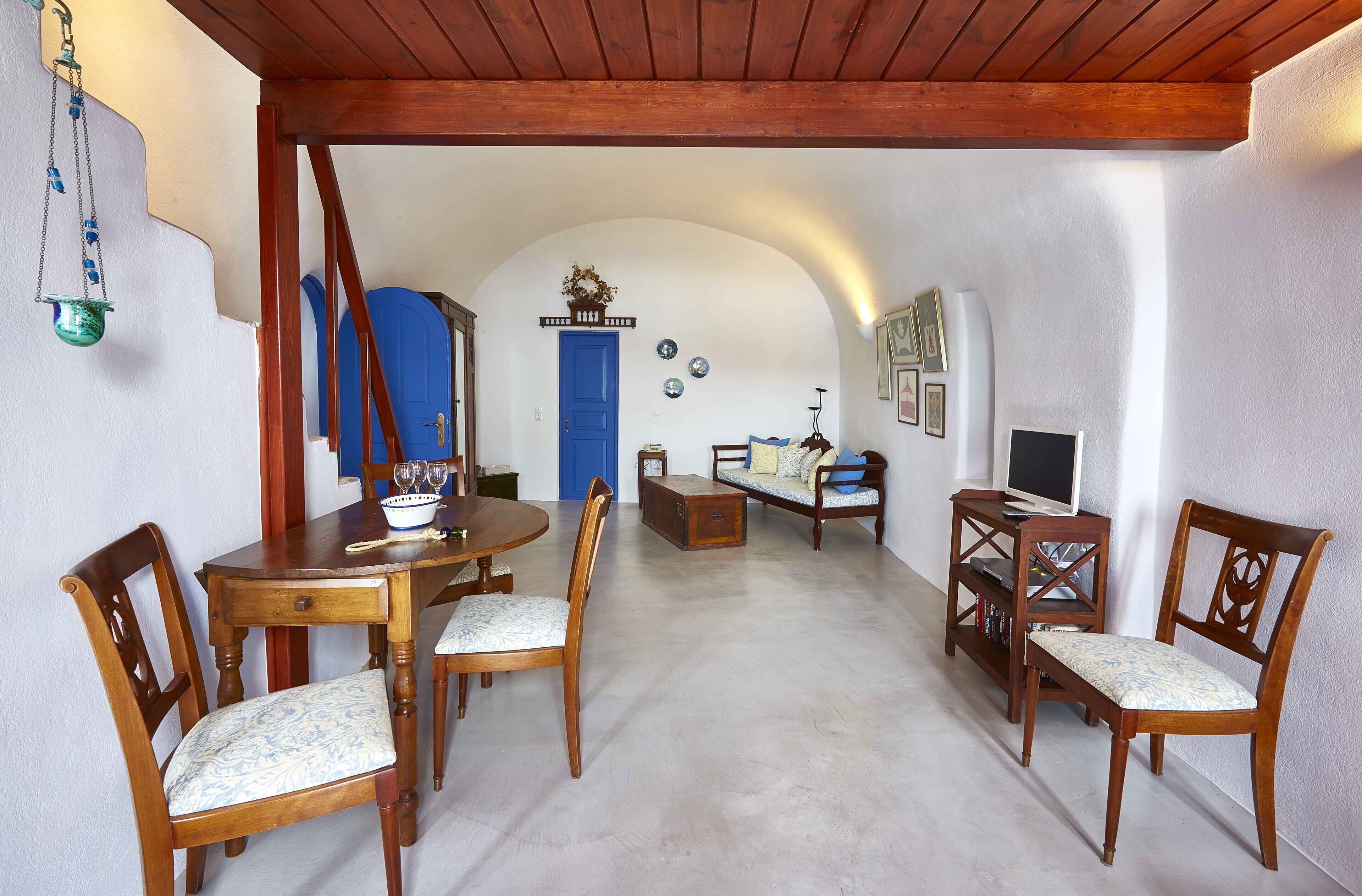 Uma área de refeições com mesa e cadeiras de madeira, uma porta azul e uma sala de estar com sofá e televisão.