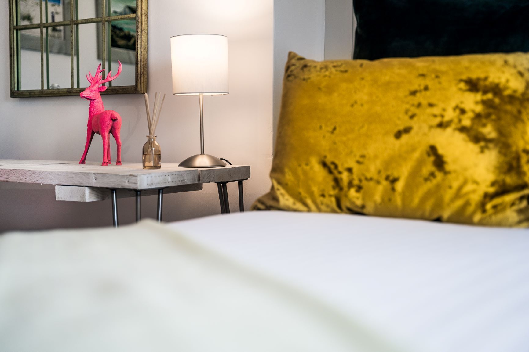Uma cama com lençol branco, uma pequena mesa de cabeceira com abajur, uma figura de cervo rosa decorativa e um travesseiro dourado.