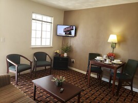 Quarto de hotel com televisão na parede, mesa de jantar com cadeiras, mesa de centro e abajur.